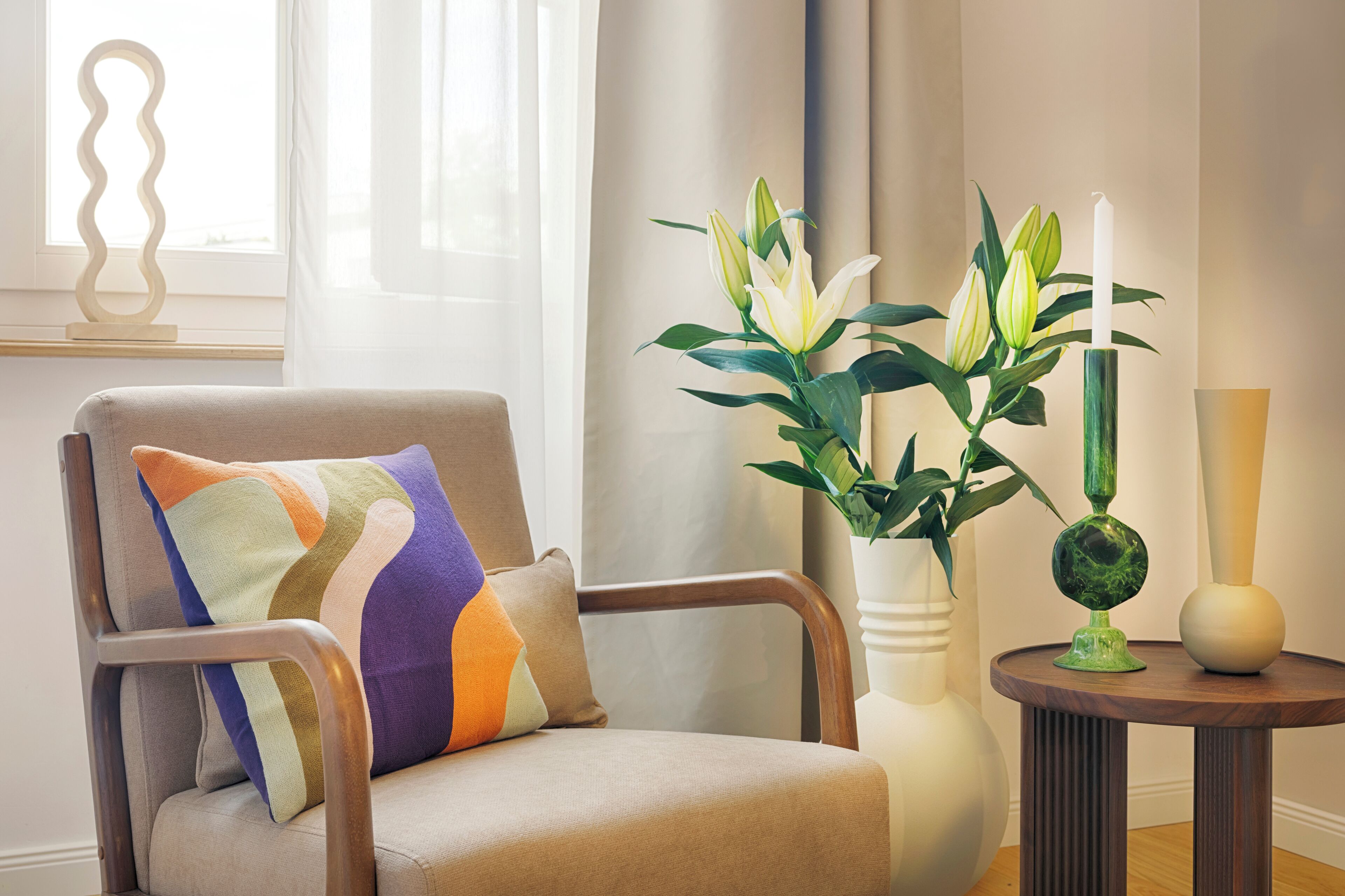 Uma sala de estar moderna com uma poltrona bege, uma almofada colorida, um vaso com flores brancas e uma pequena mesa redonda com um abajur verde.