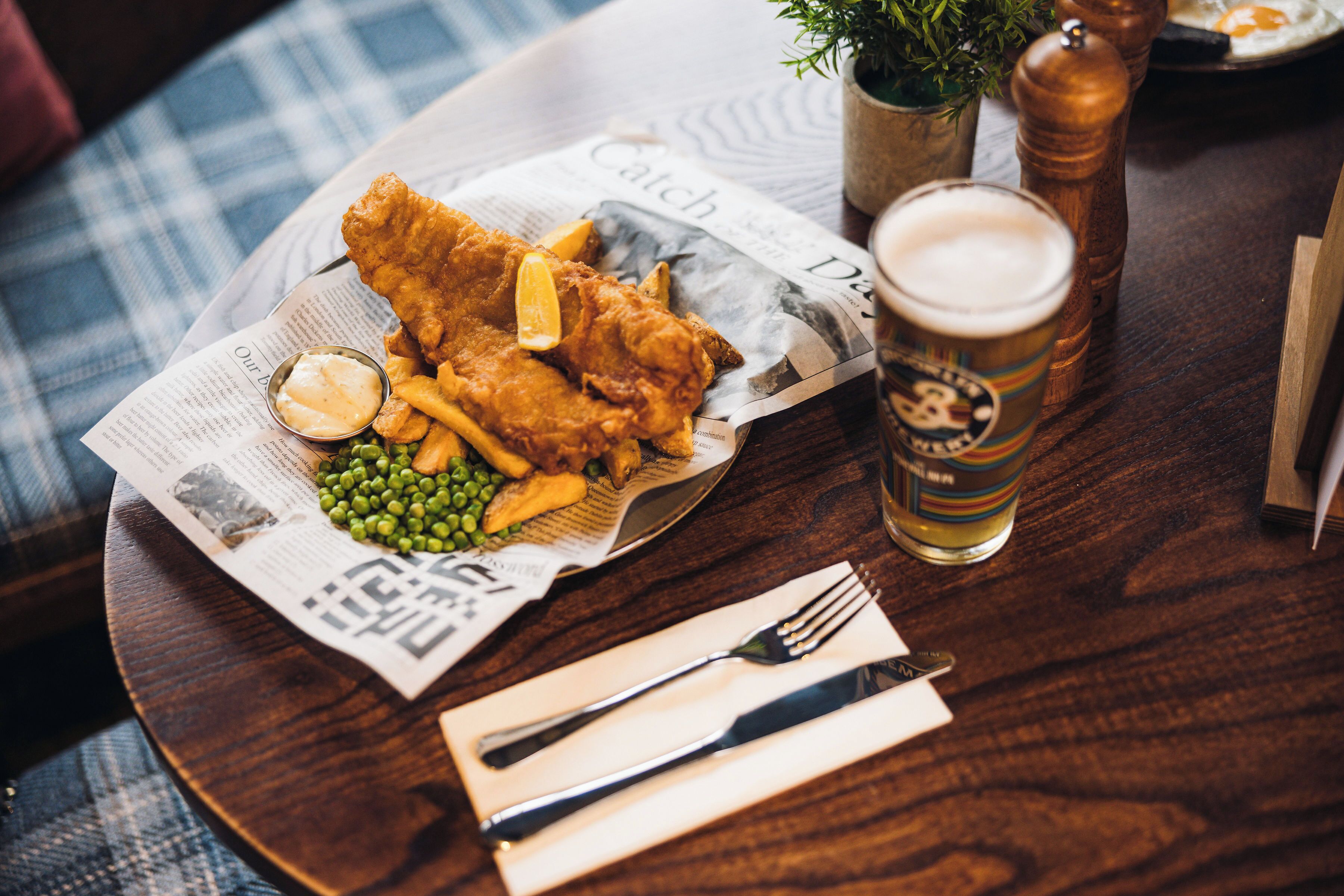 Uma refeição com peixe frito, batatas palha, ervilhas e molho sobre jornal, com um copo de cerveja e talheres numa mesa de madeira.