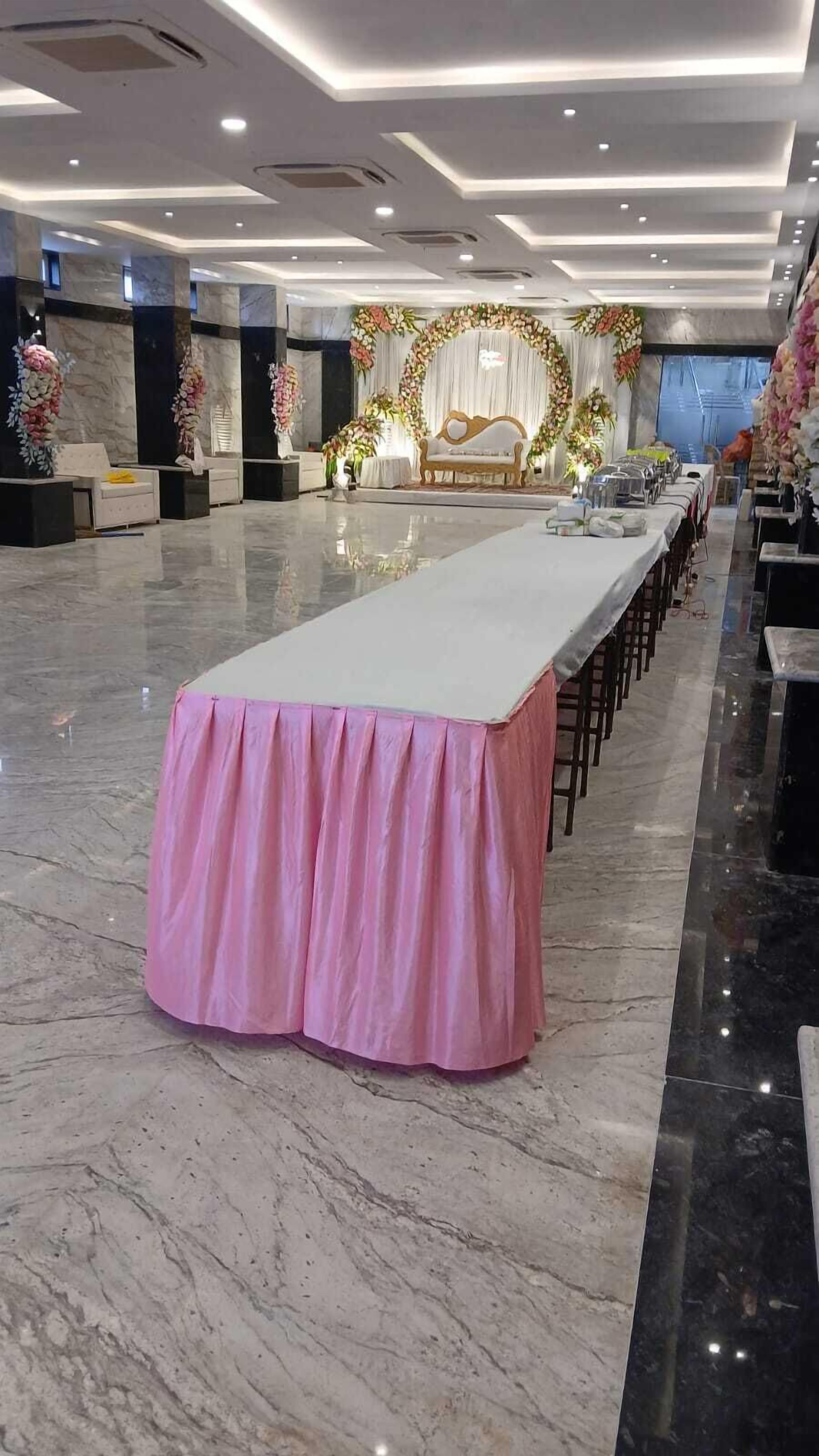 Um salão amplo com uma mesa comprida coberta por um tecido rosa, piso de mármore e um painel decorativo com arranjos florais.