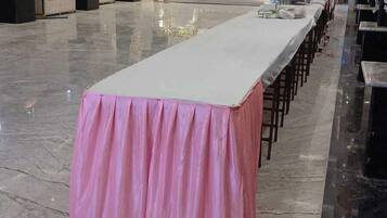 Um salão amplo com uma mesa comprida coberta por um tecido rosa, piso de mármore e um painel decorativo com arranjos florais.