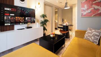 Uma sala de estar moderna com uma TV de tela plana, uma mesa de centro preta, um sofá amarelo e um espaço de jantar com mesa e cadeiras.