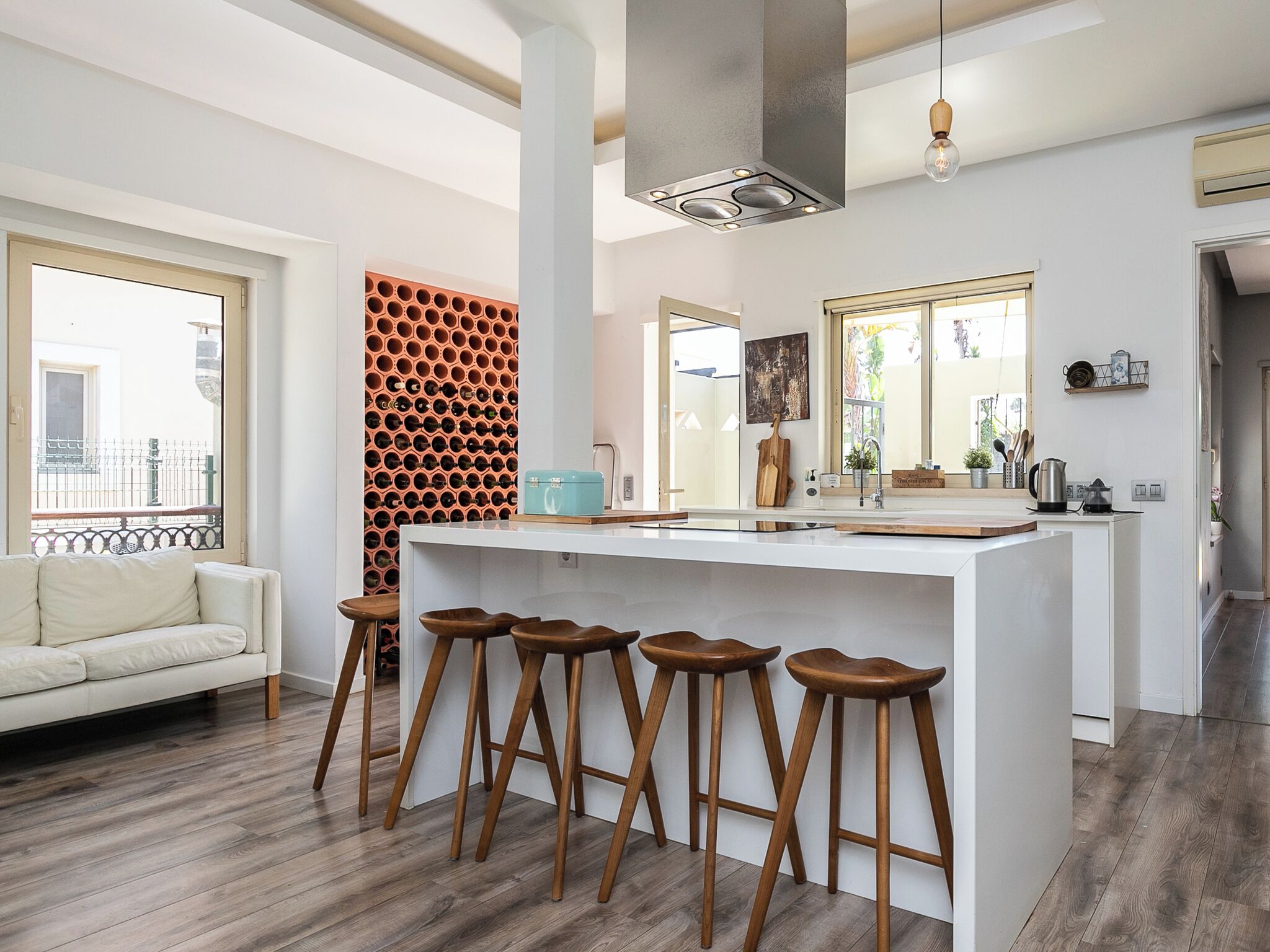 Uma cozinha moderna com uma ilha central, banquetas de madeira, espaço para vinhos e um sofá branco numa sala de estar iluminada.