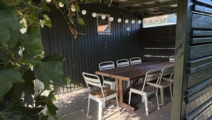 Uma área de refeições ao ar livre coberta, com uma mesa de madeira e cadeiras brancas, cercada por paredes de madeira escura e vegetação.