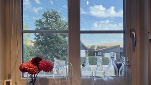 Um quarto com uma janela grande, um vaso com flores e três garrafas de vidro transparente sobre uma mesa.