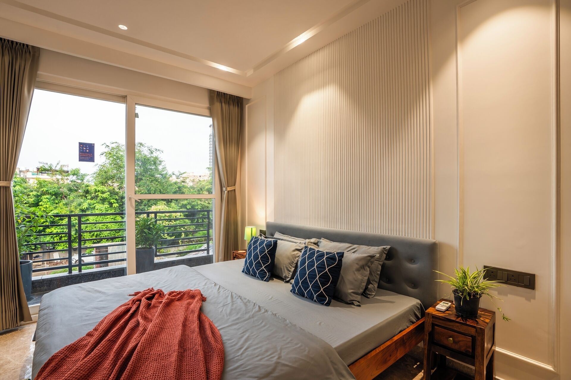 Quarto com uma cama grande, uma mesa de cabeceira, um vaso com planta e vista para o verde pela janela.