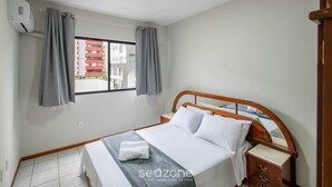 Quarto com cama, mesa de cabeceira, cômoda e janela com cortinas.