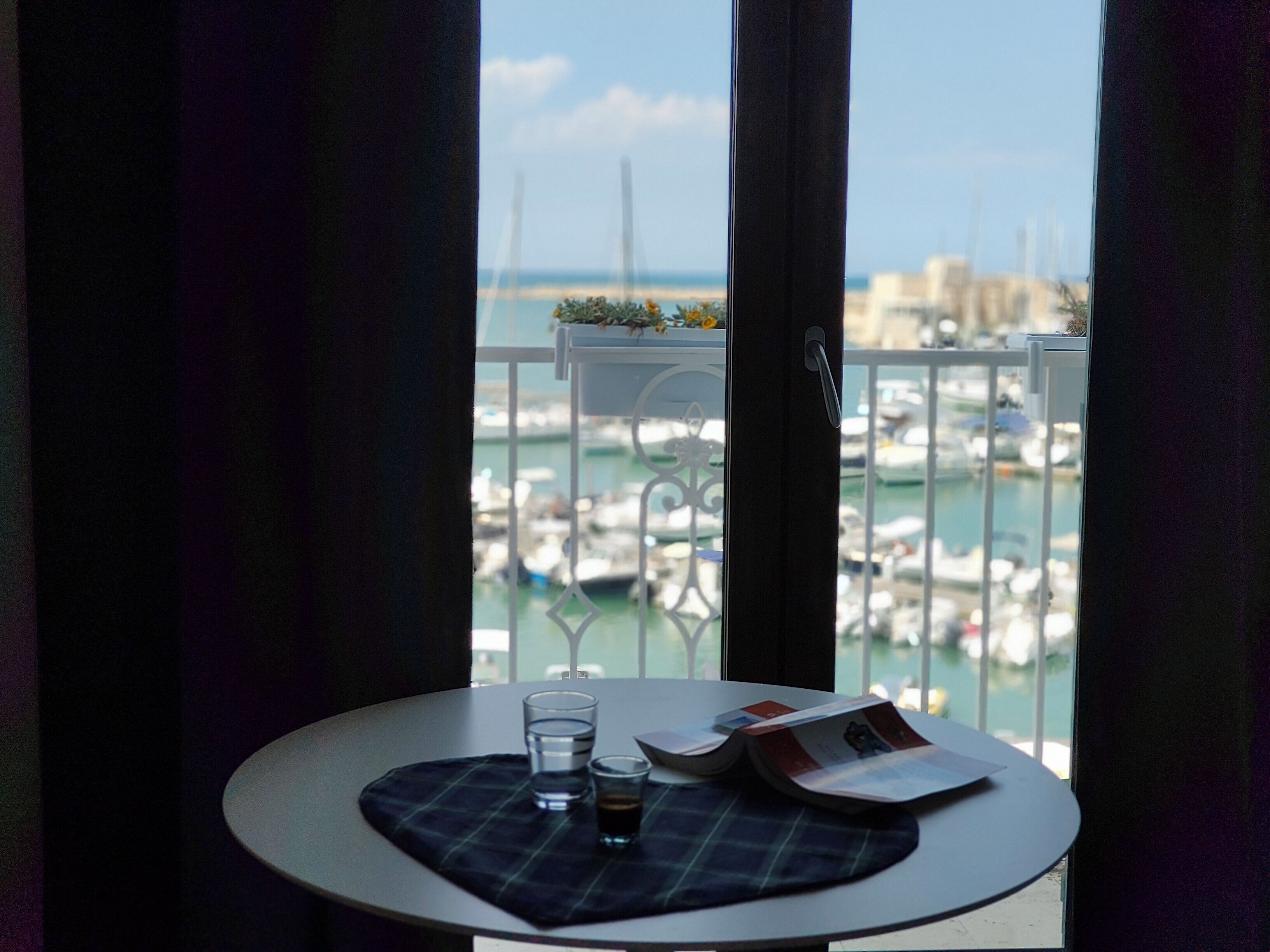 Uma mesa com um descanso para prato, um copo e um livro, com vista para uma marina com barcos e um horizonte urbano.