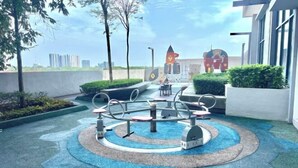 Um parquinho com equipamentos de ginástica circulares, uma pequena mesa e cadeiras, cercado por vegetação e um edifício com um mural.