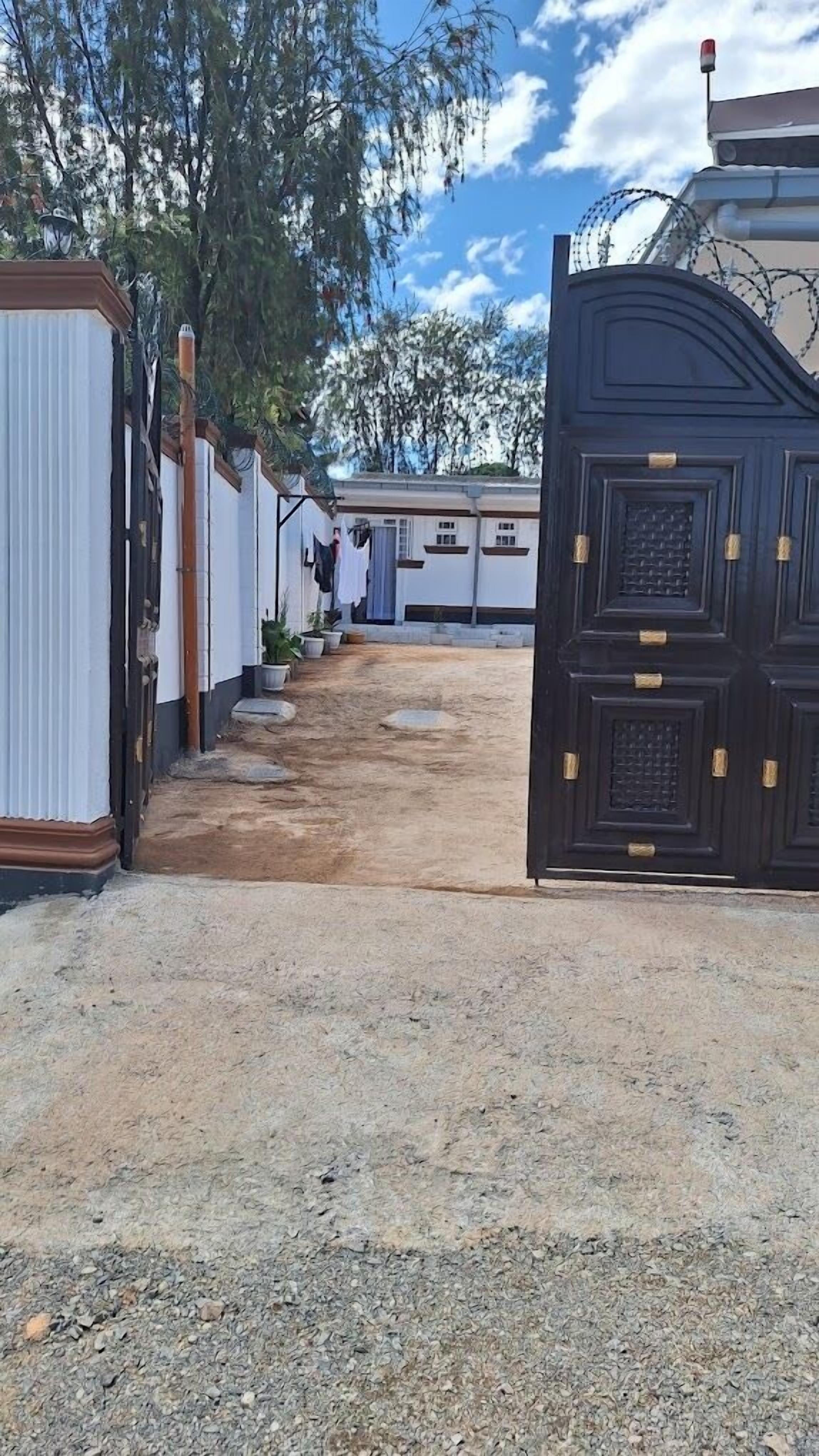 Uma entrada gradeada com um caminho de terra que leva a um edifício de paredes brancas e porta escura.