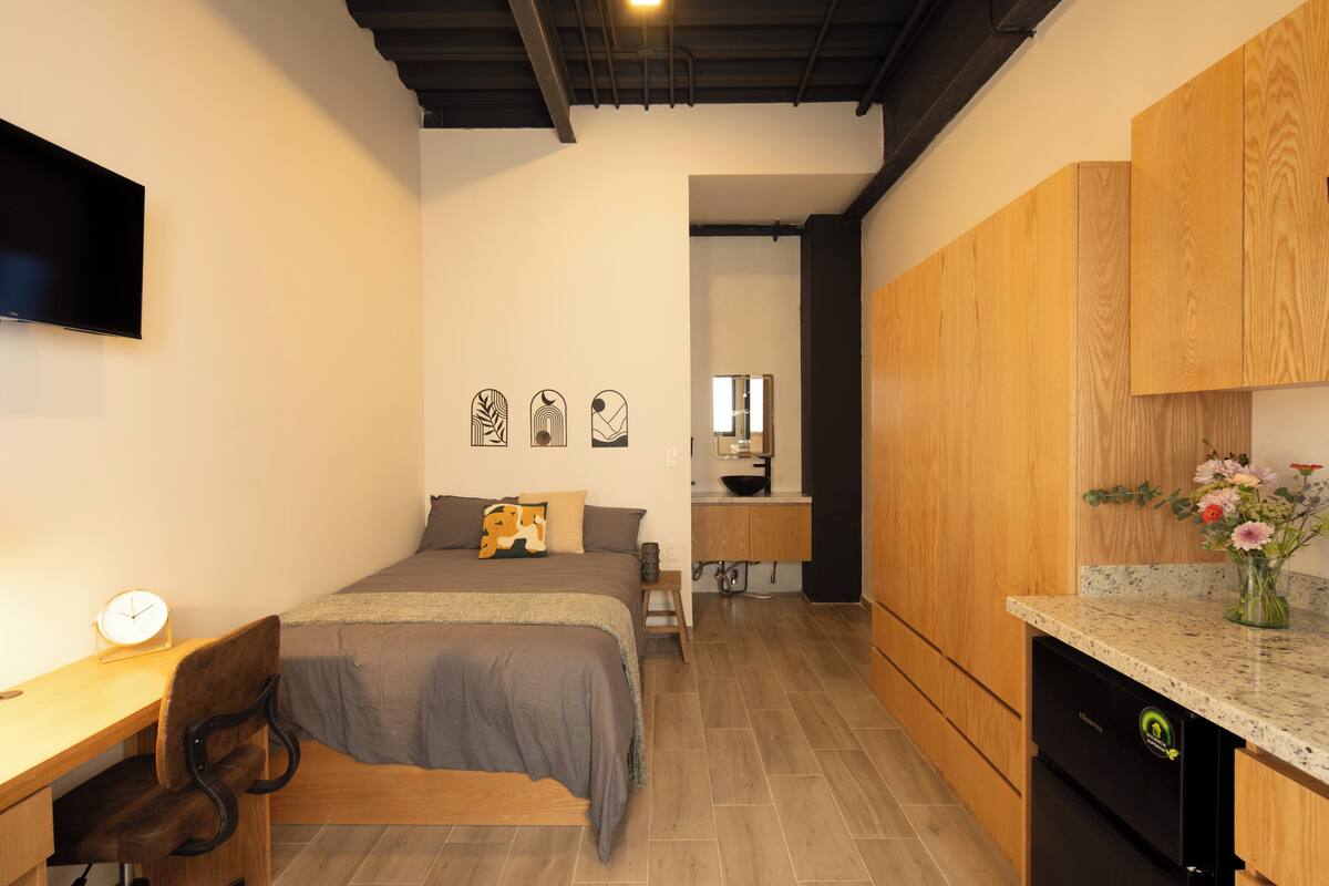 Quarto com cama, escrivaninha, cadeira, televisão e banheiro visível ao fundo.