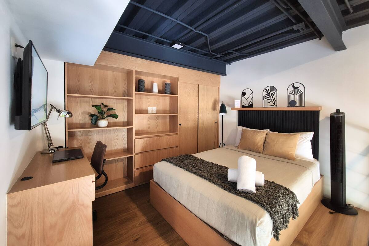 Um quarto moderno com teto estilo loft, uma cama com cabeceira, uma escrivaninha com cadeira, uma TV e uma estante com objetos decorativos.