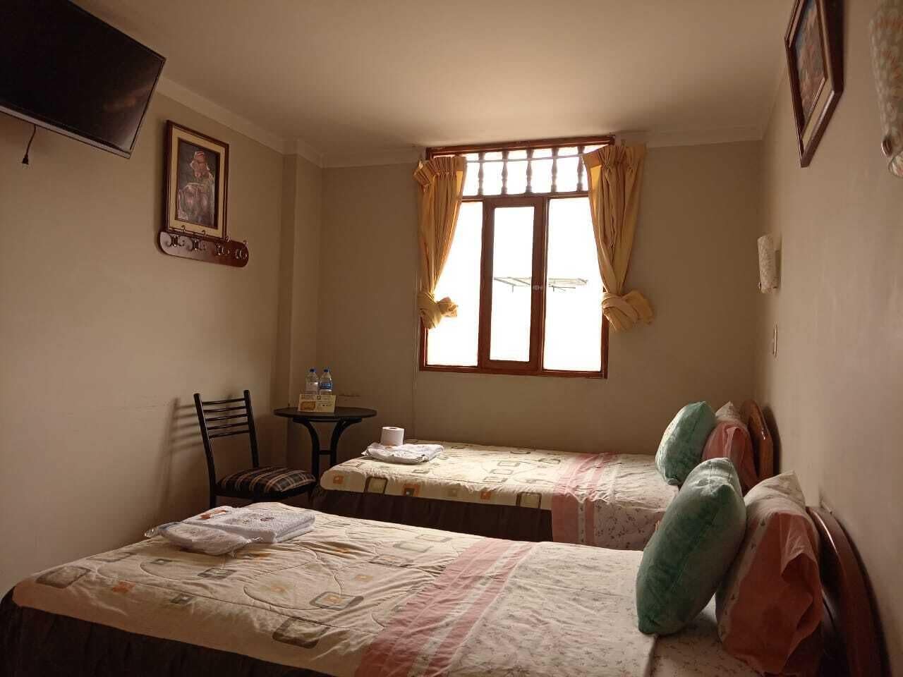 Quarto com duas camas, uma cadeira, uma mesinha e janela com cortinas.