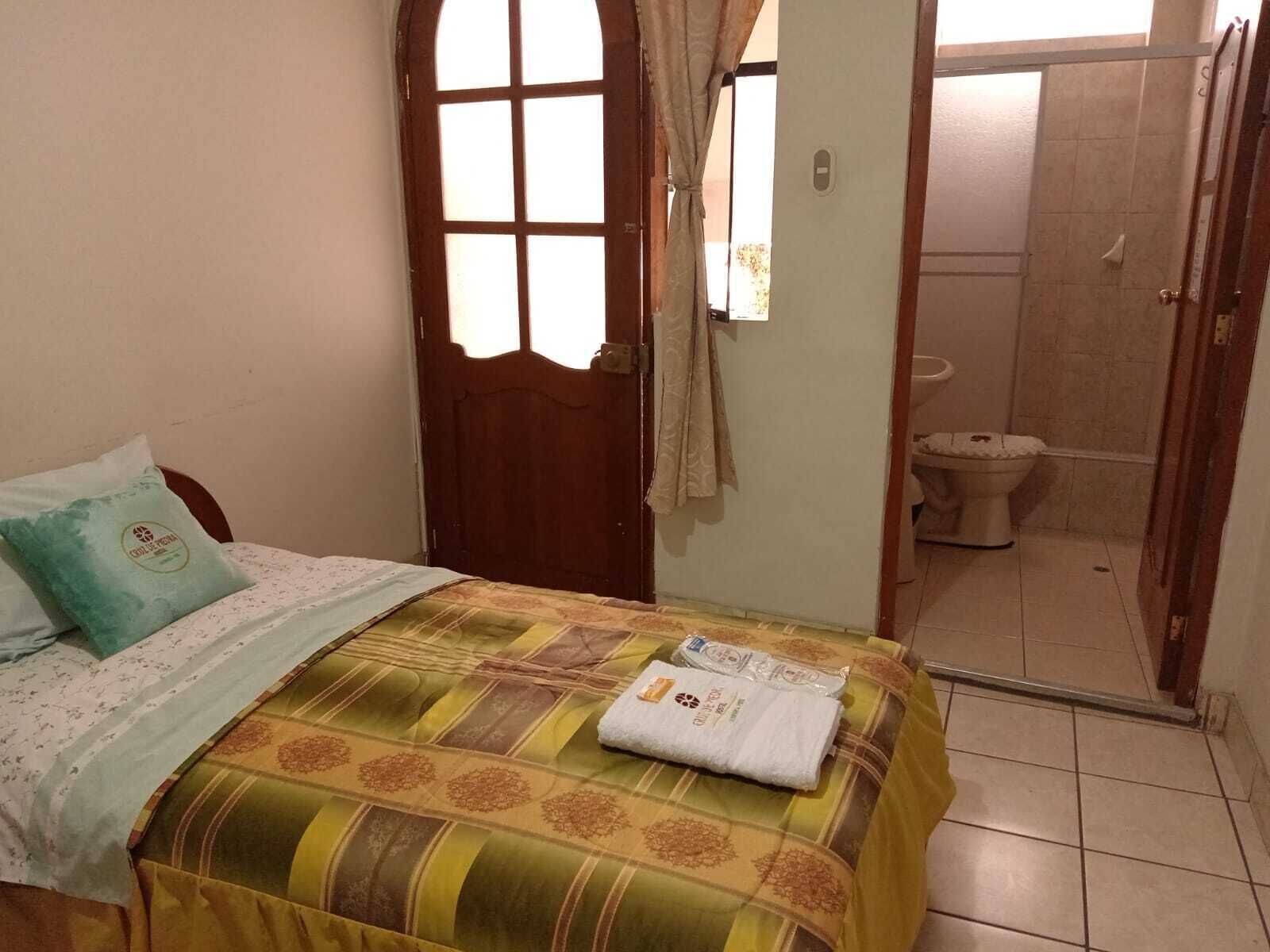 Quarto com cama, porta de madeira e banheiro visível através de uma porta aberta.