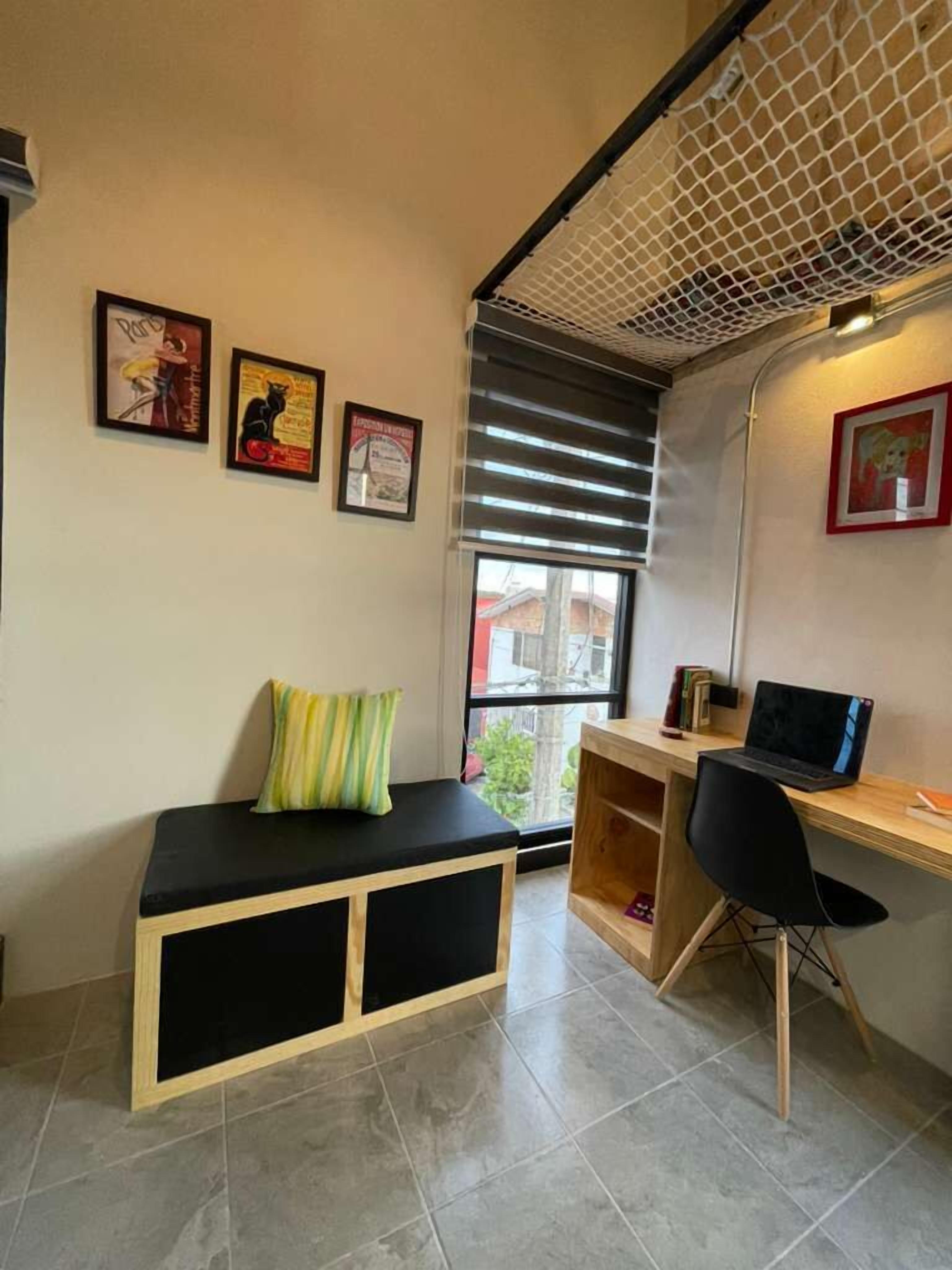 Um quarto pequeno e acolhedor, com um banco de madeira, uma escrivaninha com um laptop e uma janela com persianas.