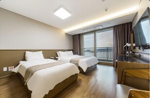 Quarto de hotel com duas camas, uma escrivaninha, uma cadeira e uma janela grande com cortinas.
