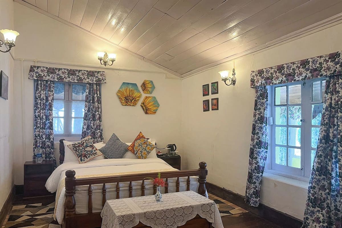 Um quarto com uma cama, uma mesinha de cabeceira, uma mesa com toalha de renda e uma janela com cortinas florais.