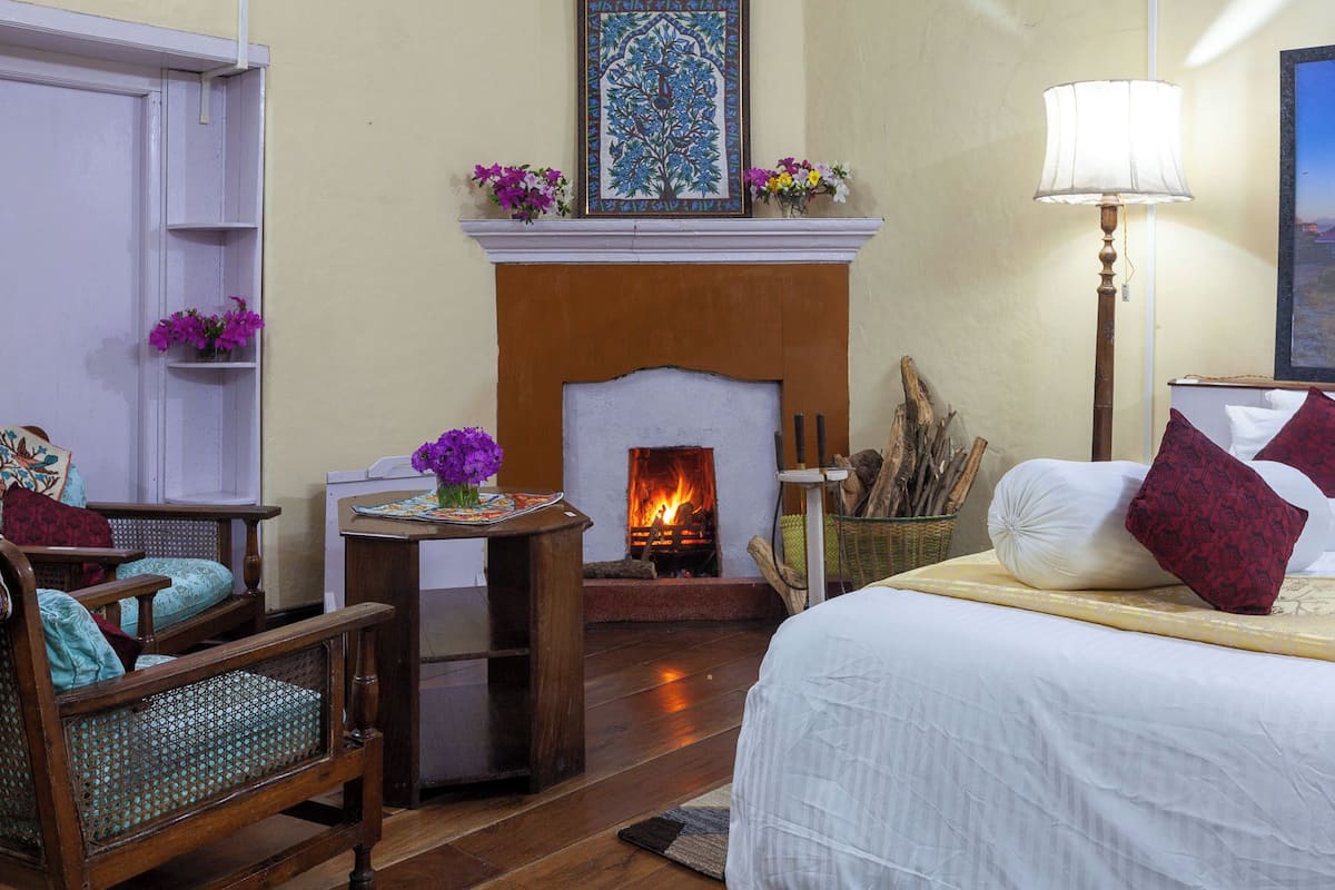 Um quarto com lareira, uma cama com almofadas, uma cadeira, uma mesa com flores e um abajur.