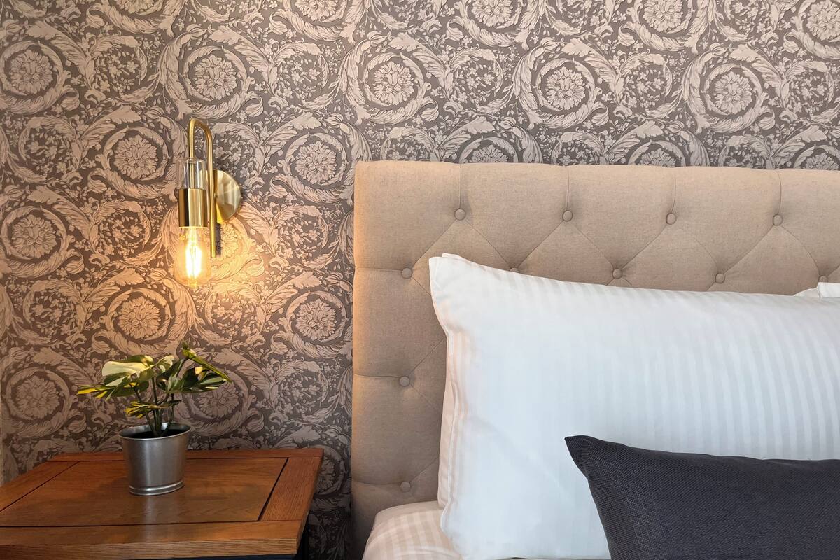 Quarto com cabeceira estofada, mesa de cabeceira com uma planta e papel de parede estampado.