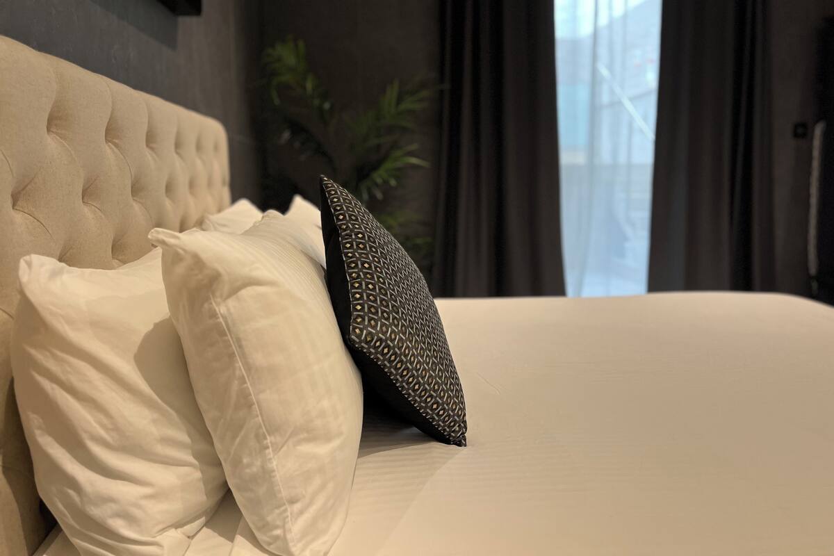 Uma cama com almofadas brancas e estampadas, cabeceira com detalhe em matelassê e vista de um edifício pela janela.