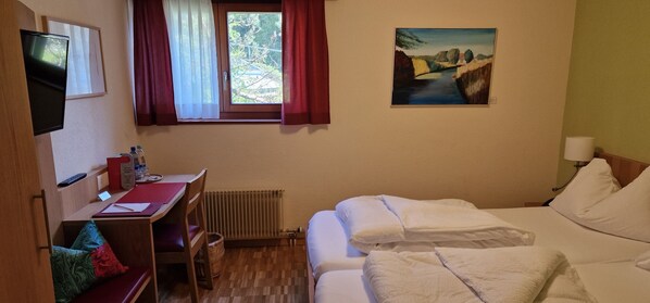 Quarto de hotel com cama, escrivaninha com cadeira, televisão, janela com cortinas e um quadro na parede.