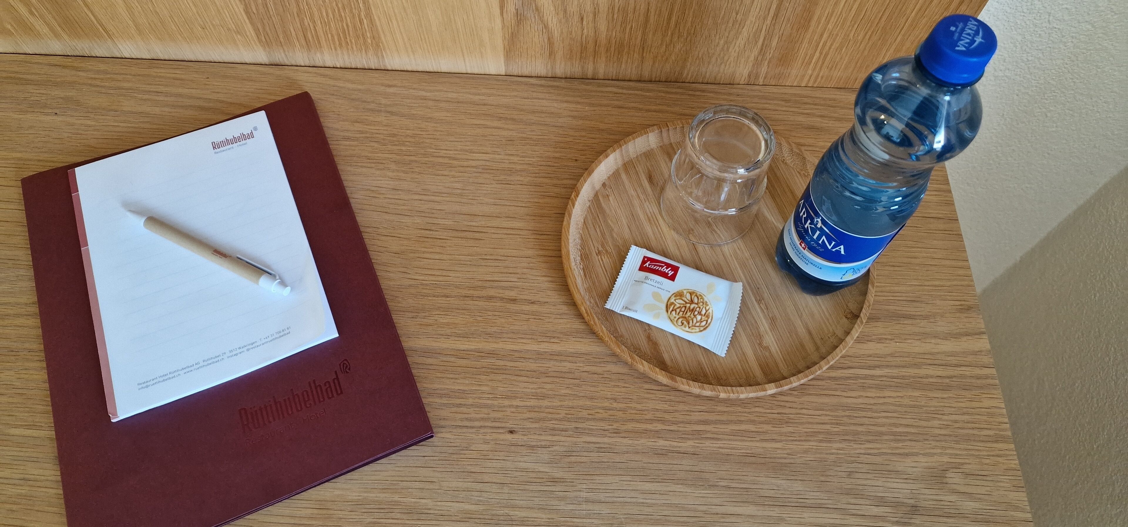 Uma mesa de madeira com um caderno vermelho, uma caneta, um copo, uma garrafa de água e um pacote de lanches.