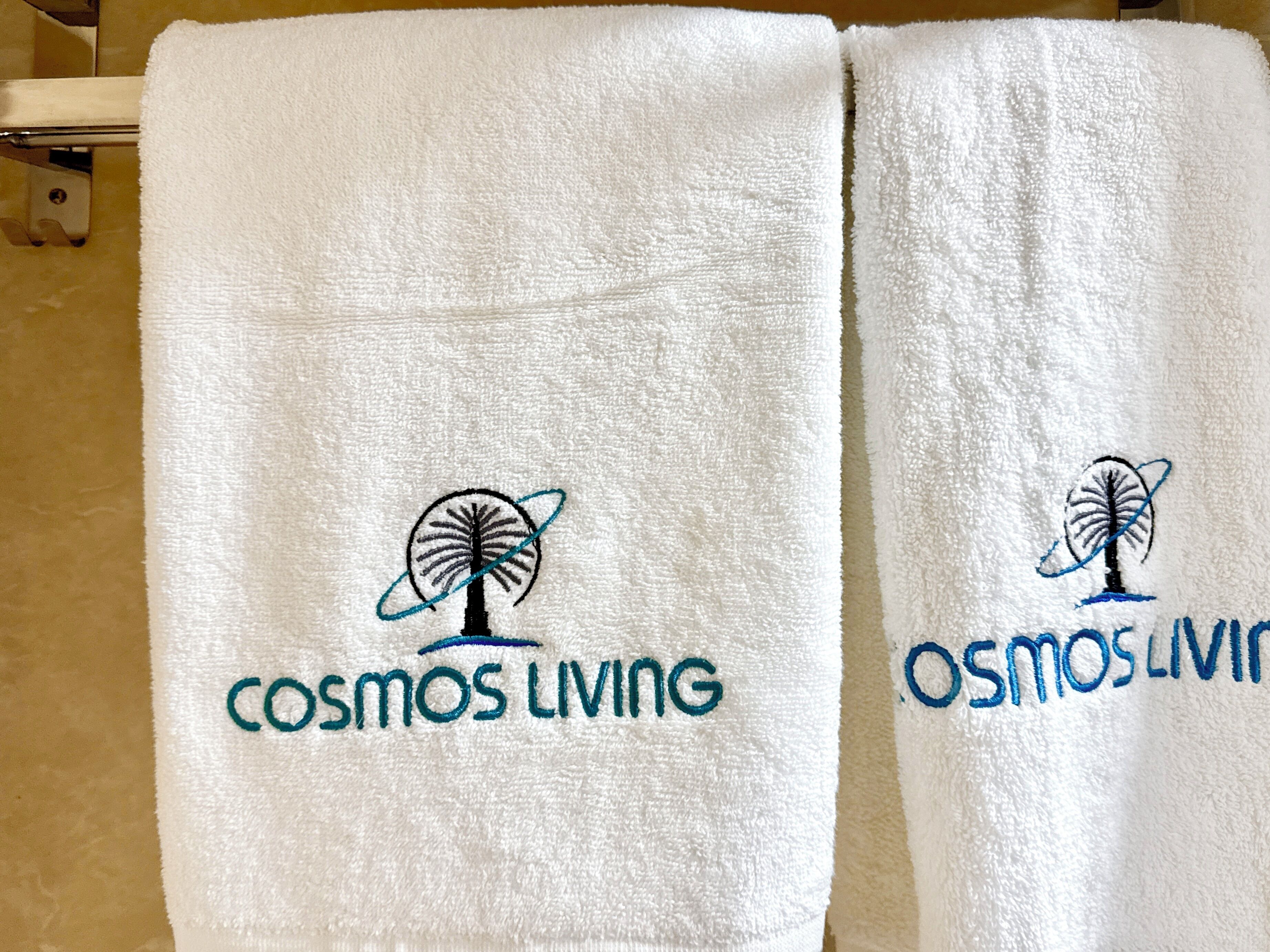 Duas toalhas brancas com logotipos e texto bordados, penduradas em um suporte de metal.