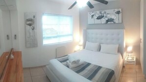 Quarto com uma cama grande, mesa de cabeceira com abajur, janela com persianas e obras de arte nas paredes.