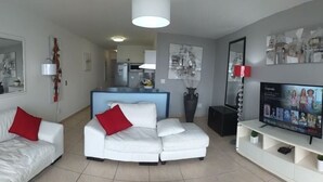 Uma sala de estar moderna com um sofá branco, uma TV de tela plana e uma cozinha ao fundo.