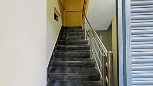 Uma escadaria com degraus pretos e corrimãos de metal, levando a uma porta com persianas horizontais.