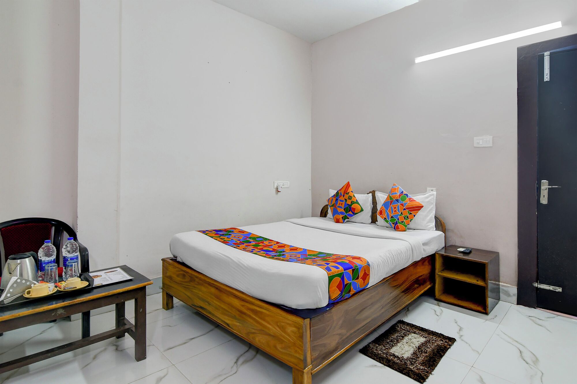 Quarto com cama de madeira, roupa de cama branca, almofadas coloridas, uma pequena mesa de cabeceira, uma cadeira e uma cafeteira.