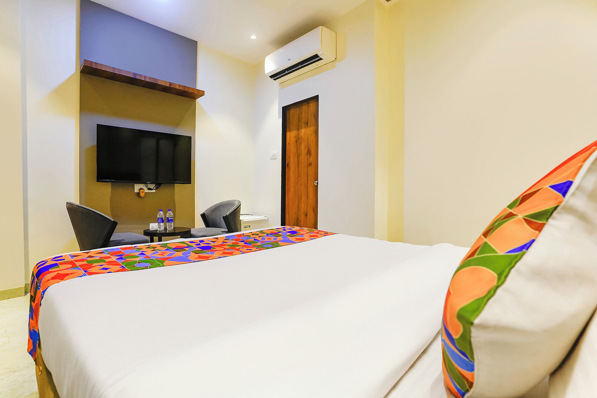 Quarto de hotel com uma cama, televisão fixada na parede, escrivaninha com duas cadeiras e ar condicionado.