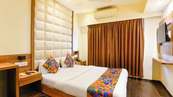 Quarto de hotel com uma cama grande, mesa de cabeceira de madeira, escrivaninha, ventilador de teto, aparelho de ar condicionado e televisão na parede.