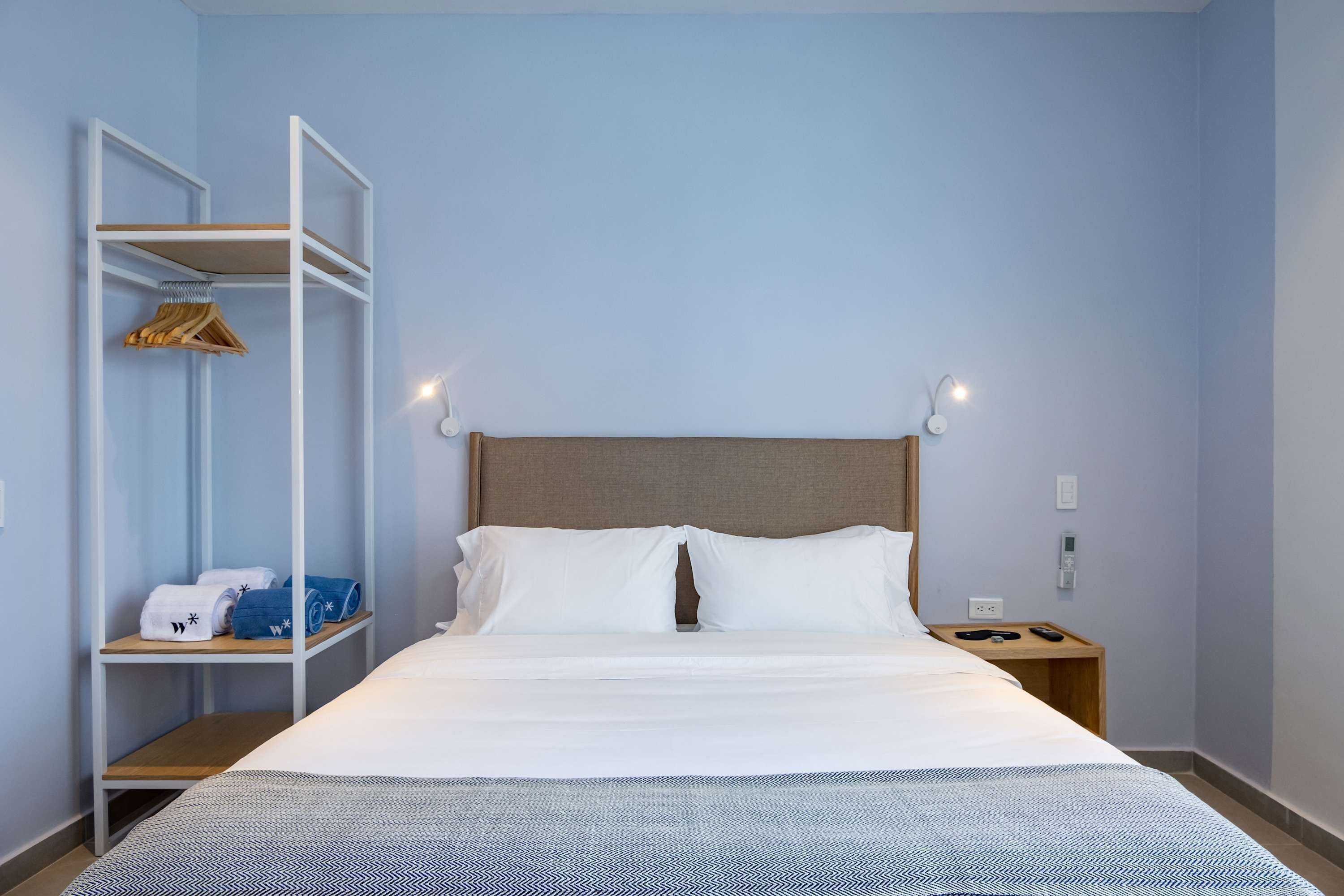 Uma cama bem arrumada com lençóis brancos e manta estampada, uma mesa de cabeceira de madeira com abajur, e um guarda-roupa embutido com roupas penduradas.