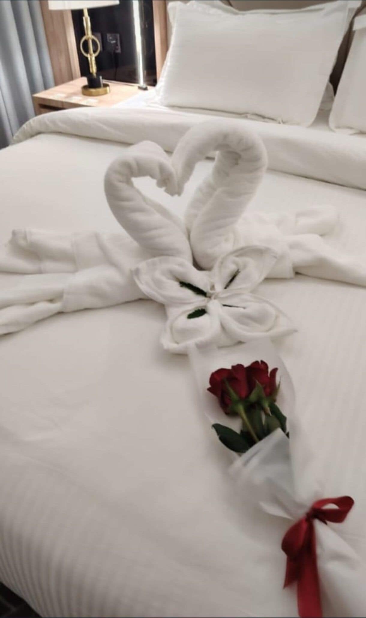 Cama bem arrumada com uma toalha disposta em formato de coração e um buquê de rosas vermelhas amarradas com um laço vermelho.