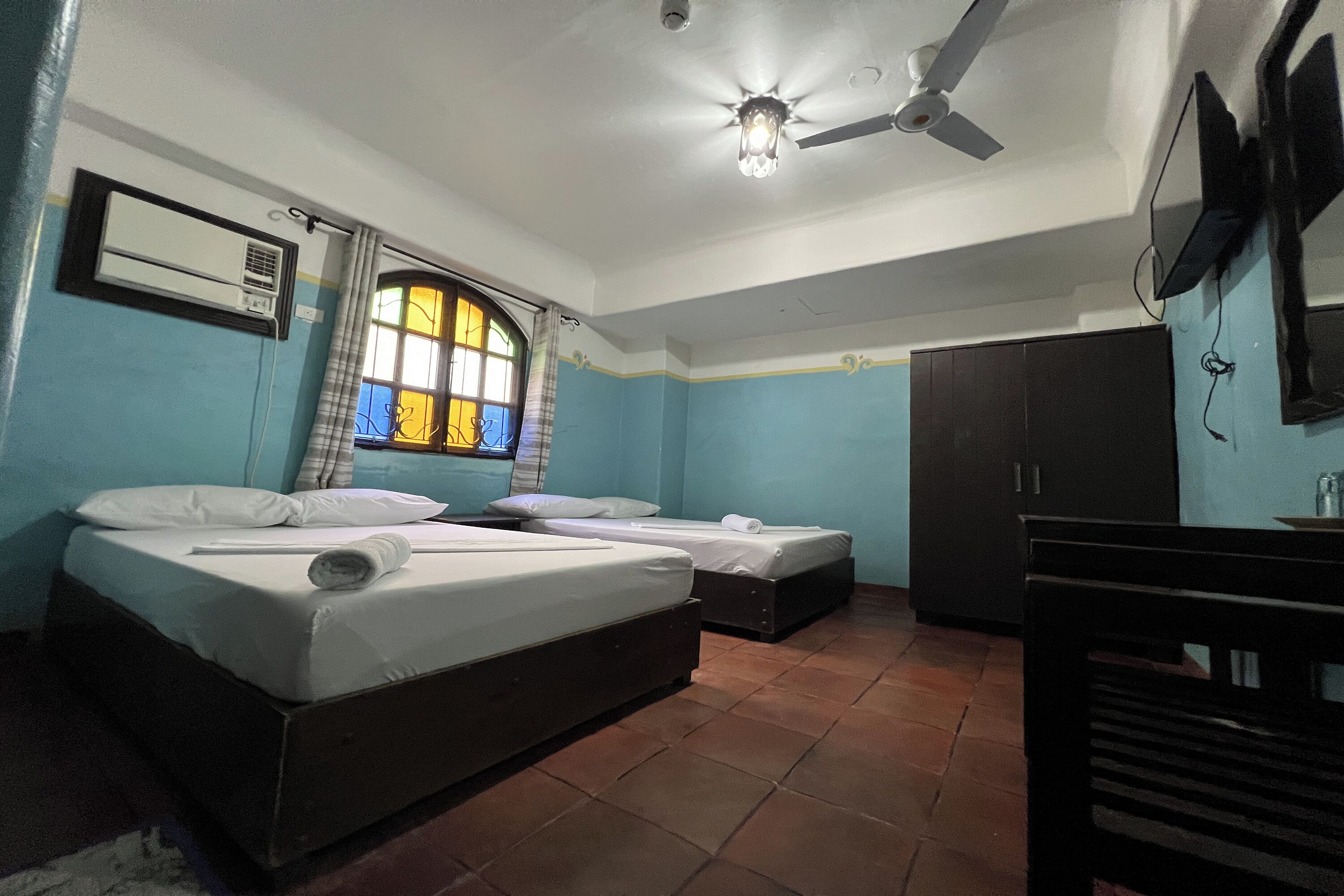 Quarto com duas camas, ventilador de teto, ar condicionado, janela com cortinas e guarda-roupa escuro.