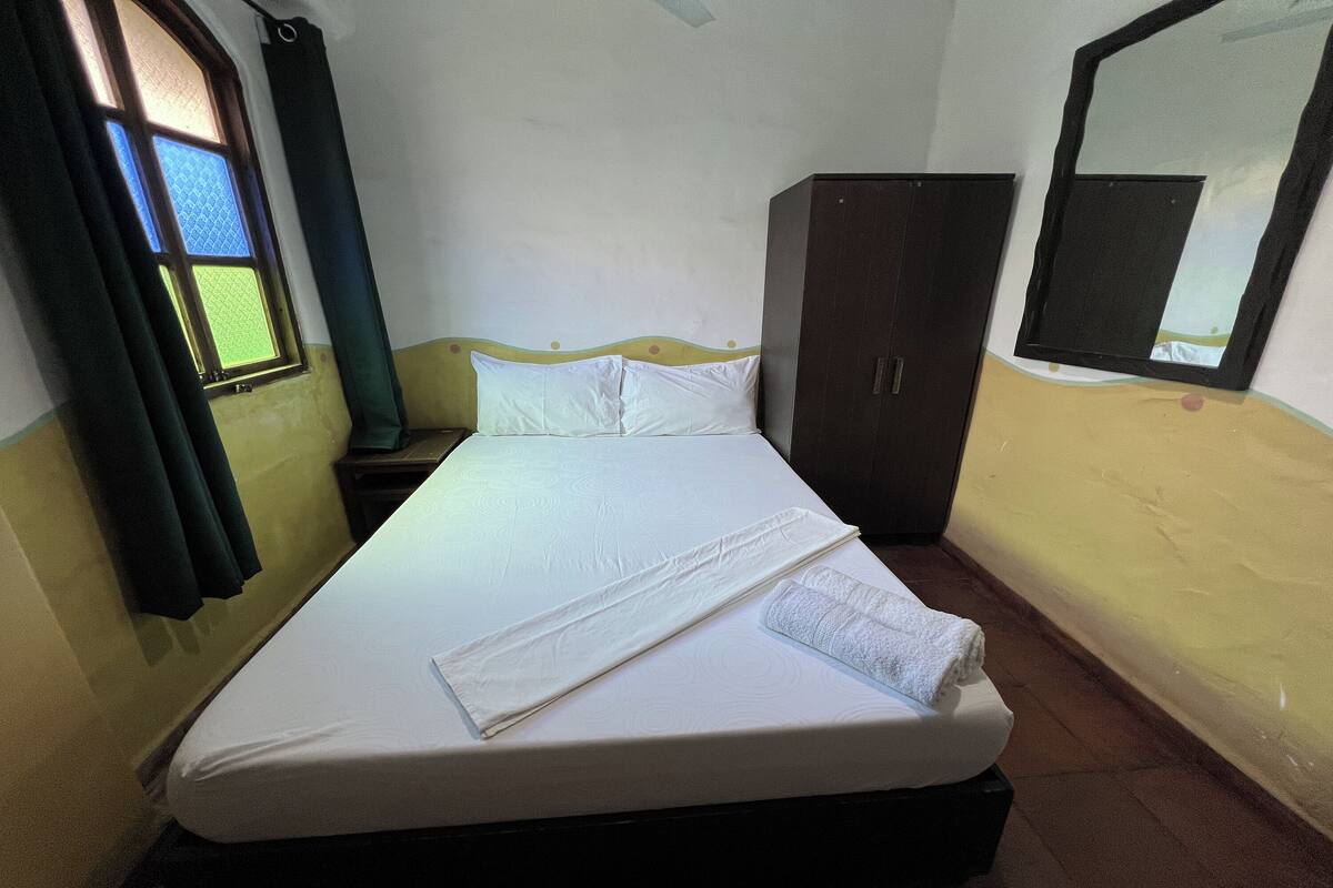 Uma cama de solteiro com lençóis brancos, um guarda-roupa de madeira escura, uma pequena mesa de cabeceira e uma janela com cortinas.