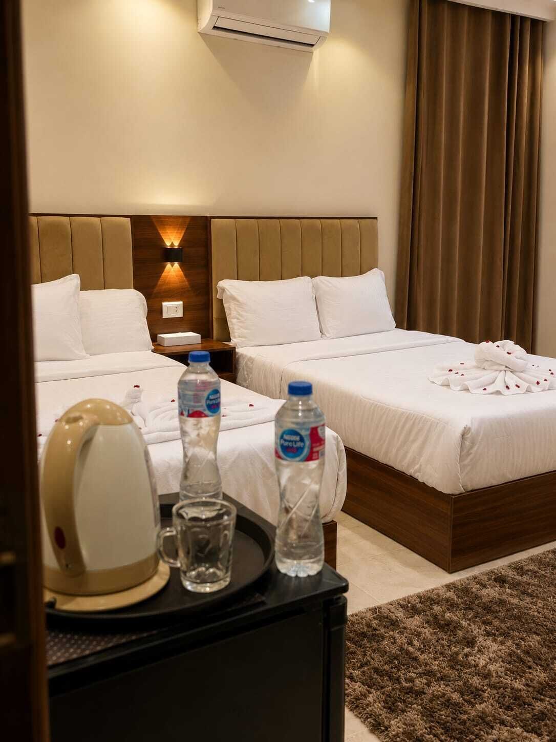 Quarto de hotel com duas camas, uma mesinha de cabeceira com chaleira, garrafas de água e um suporte para bandeja.