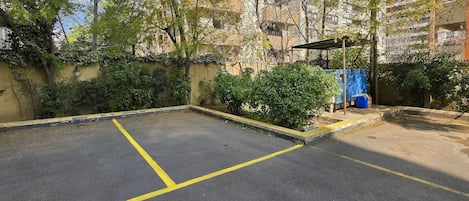 Uma área de estacionamento com linhas amarelas delimitando as vagas, rodeada por vegetação e um edifício ao fundo.