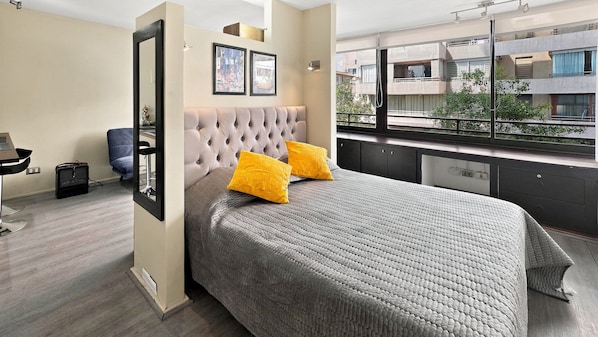 Quarto moderno com cabeceira estofada, cama grande com colcha cinza texturizada, duas almofadas amarelas vibrantes e vista da cidade através de uma janela ampla.