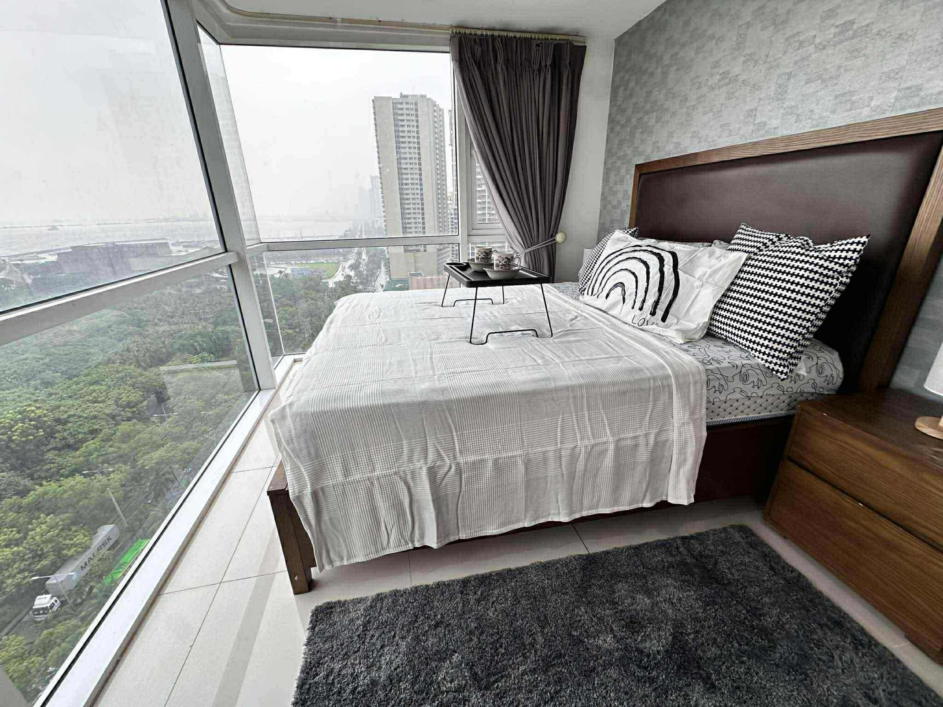 Quarto com uma janela grande, cama com colcha branca, mesa de cabeceira de madeira e tapete preto.