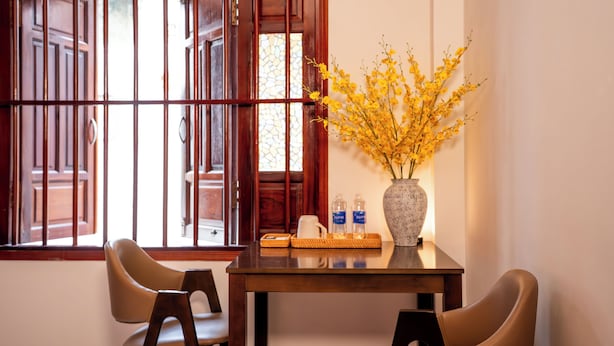 Um quarto com uma mesa de madeira, duas cadeiras, um vaso com flores amarelas e uma janela com grades de metal.