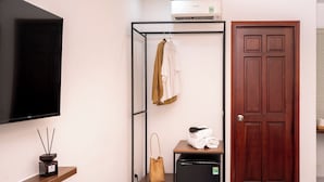 Quarto de hotel com prateleira de madeira, uma pequena geladeira preta, cabideiro, porta marrom e televisão fixada na parede.