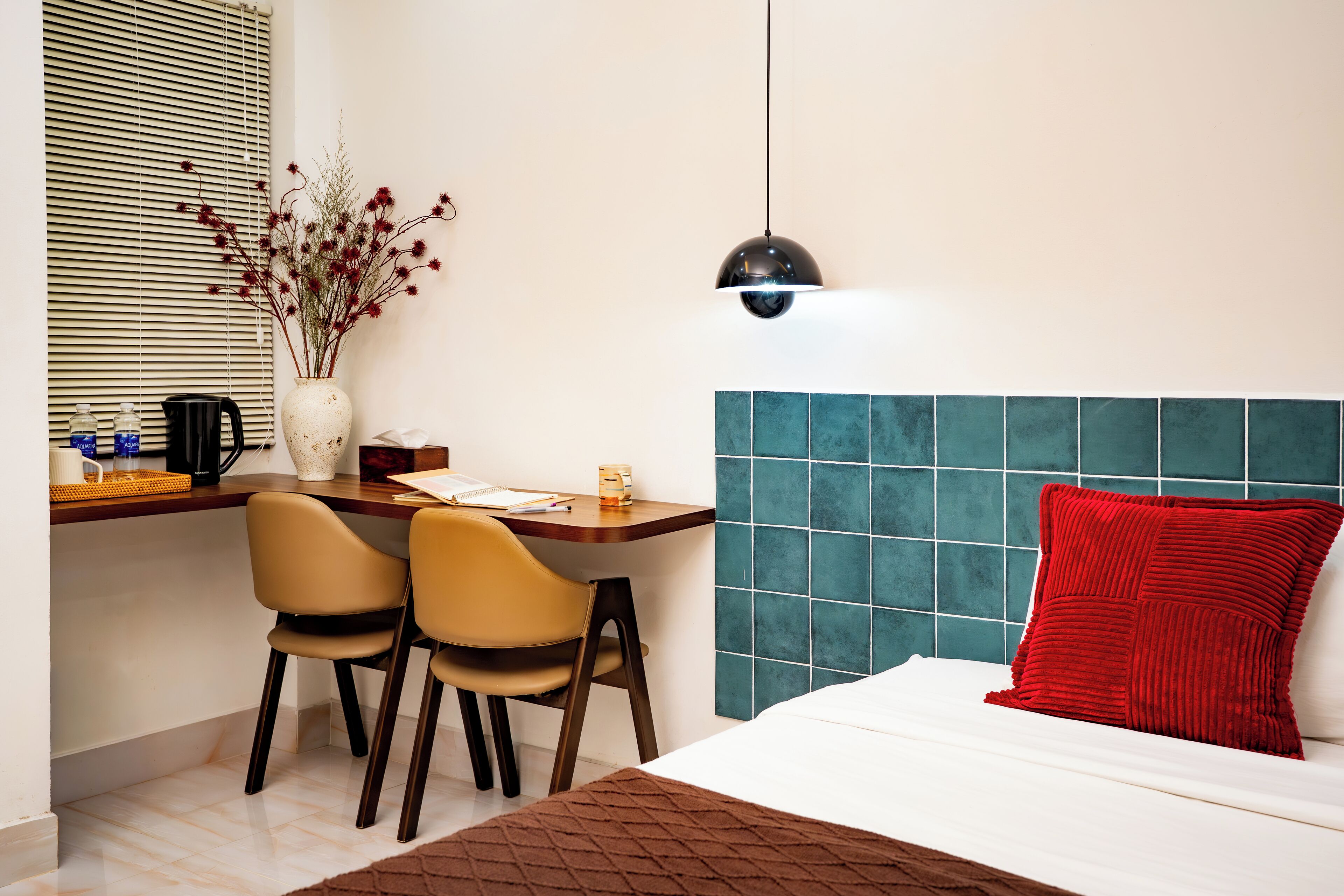 Quarto de hotel com cama, escrivaninha com cadeiras, vaso com flores, cafeteira e luminária pendente.