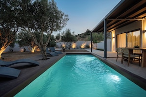 Uma piscina com espreguiçadeiras e uma mesa, cercada por árvores e um edifício com janelas iluminadas.