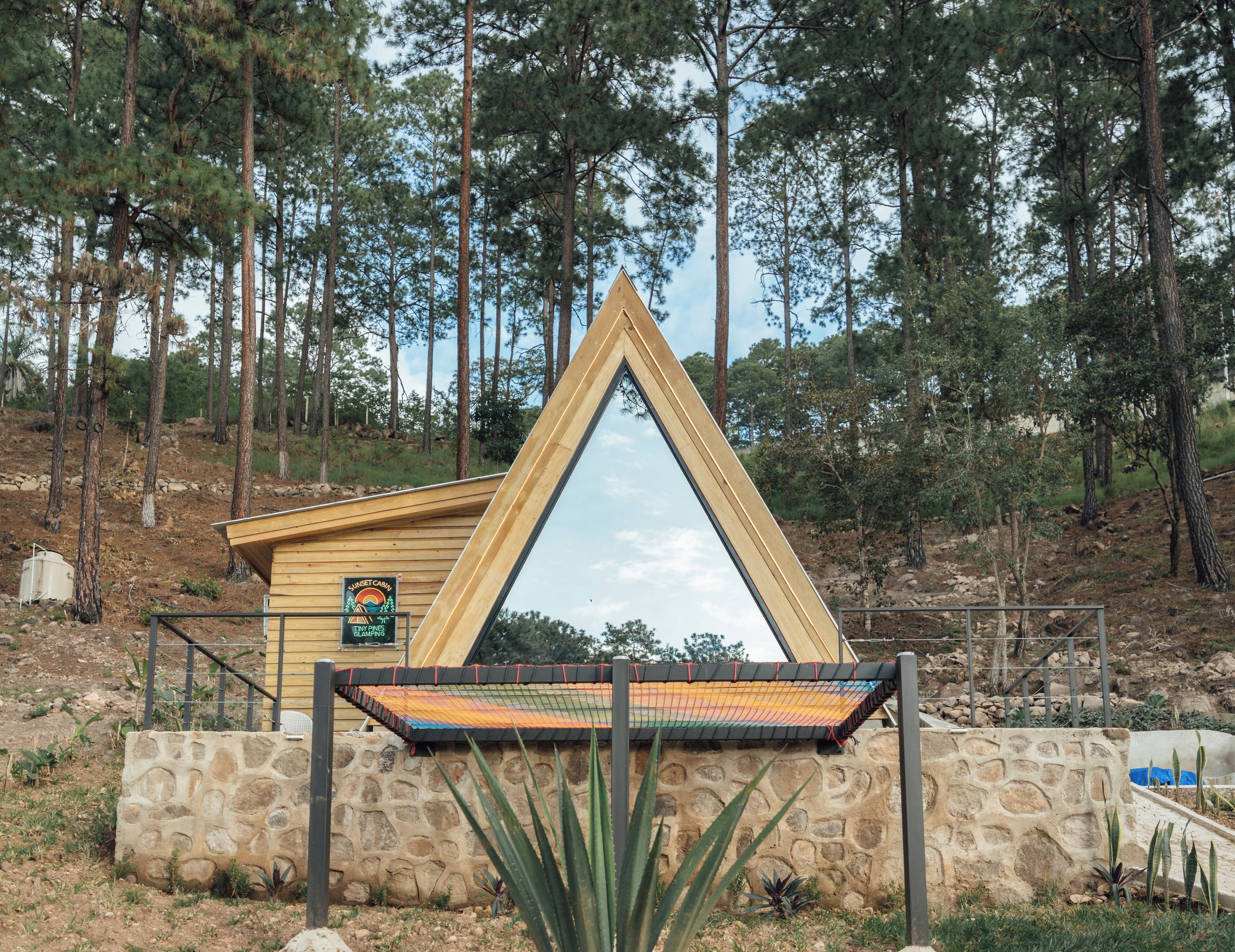 Estrutura de madeira triangular com uma placa indicando Sunset Cabin, cercada por um muro de pedra e vegetação.