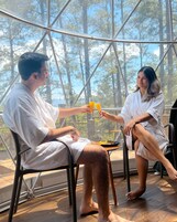Duas pessoas sentadas à mesa dentro de um espaço com vista para árvores.