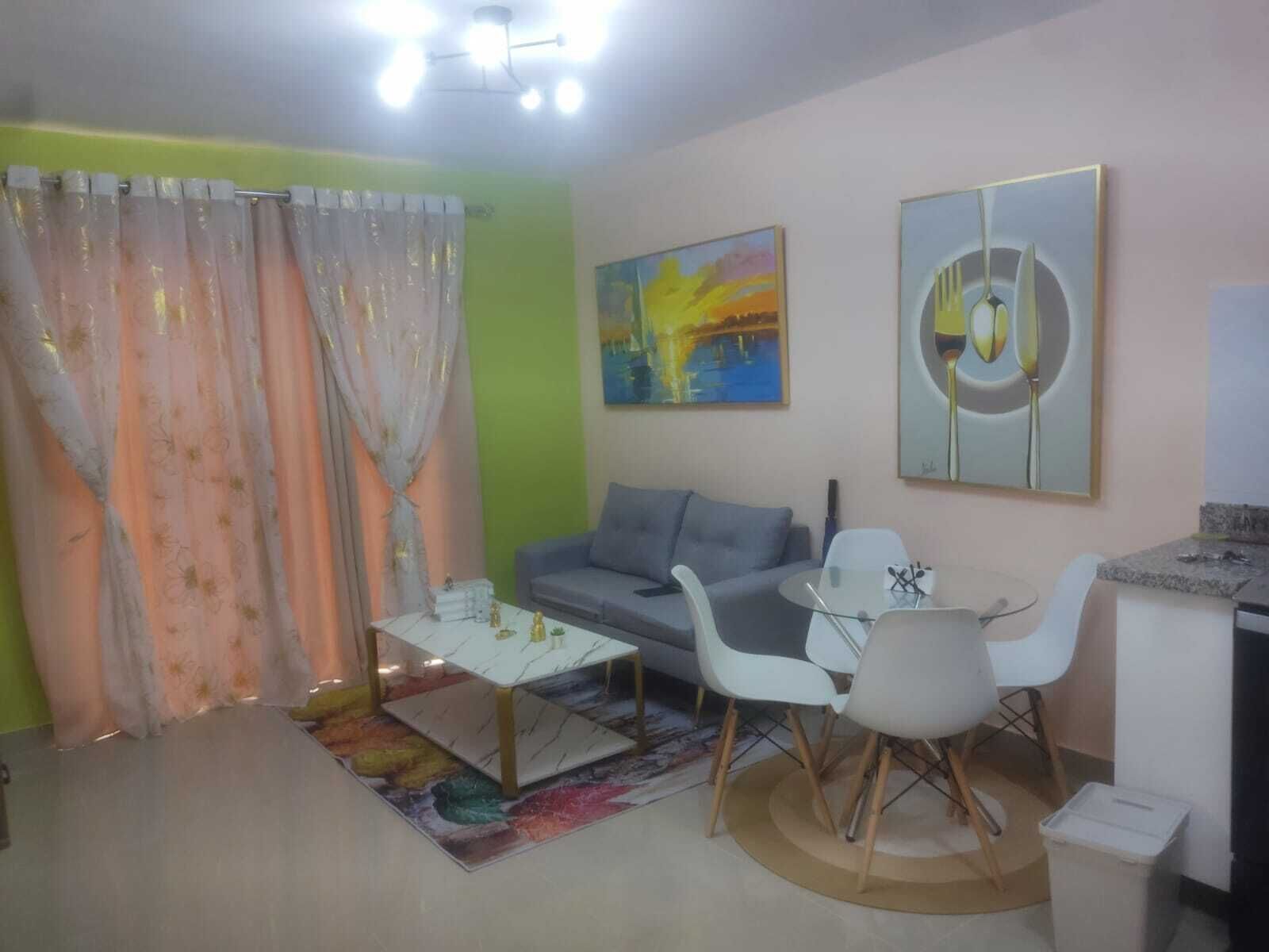 Uma sala de estar com sofá, mesa de centro e cadeiras, uma área de jantar com mesa e cadeiras, e duas pinturas nas paredes.
