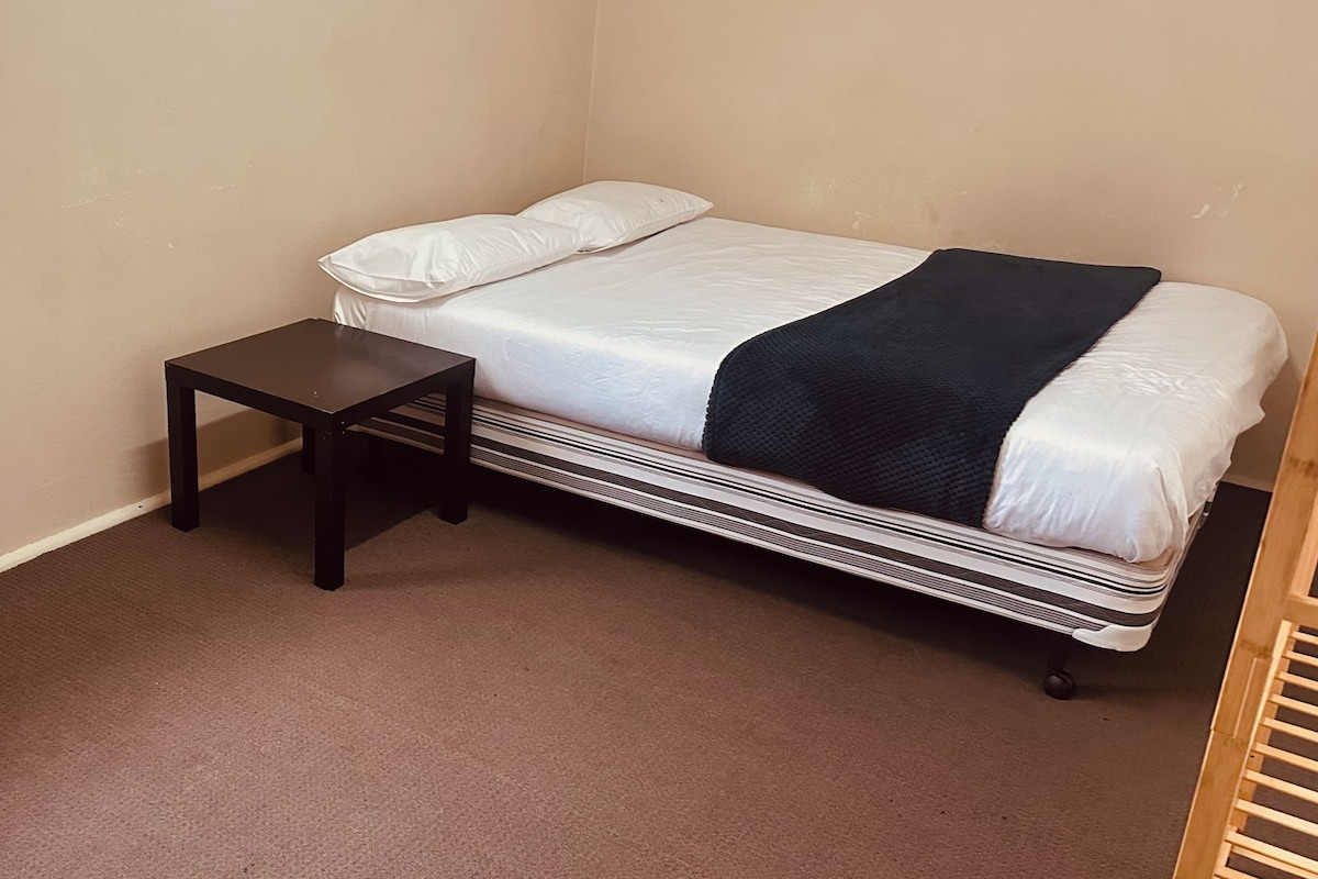 Uma cama de solteiro com colchão branco e manta escura, uma mesinha de cabeceira preta e uma escada de madeira encostada na parede.