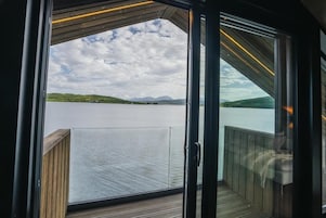 Vista de um quarto com porta de vidro que dá para uma varanda com vista para um lago e colinas ao longe.