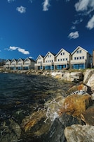 Uma fileira de casas à beira-mar, com varandas, localizada em uma orla rochosa com águas azuis cristalinas.
